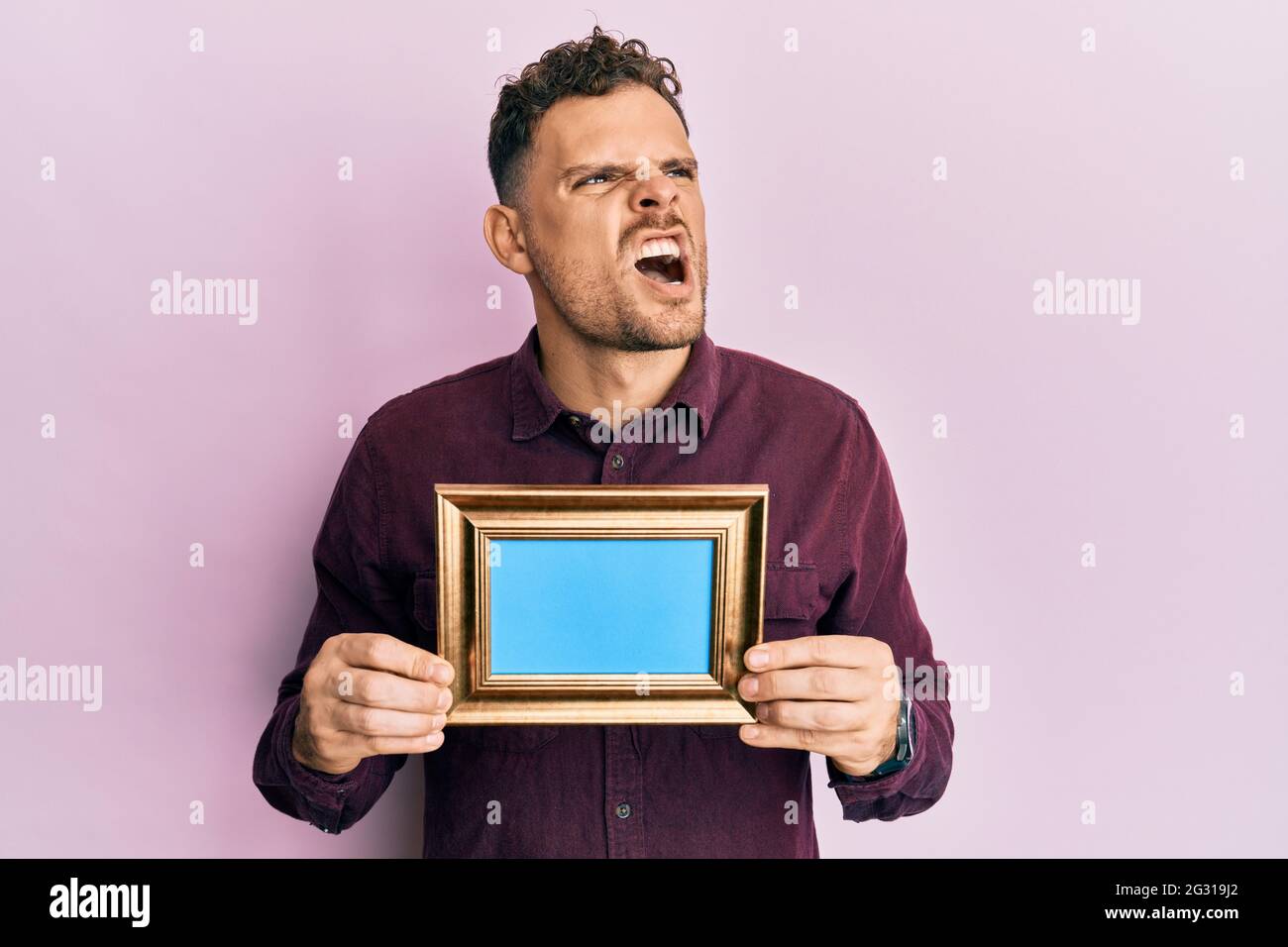 Young hispanic man holding empty frame angry and mad screaming ...
