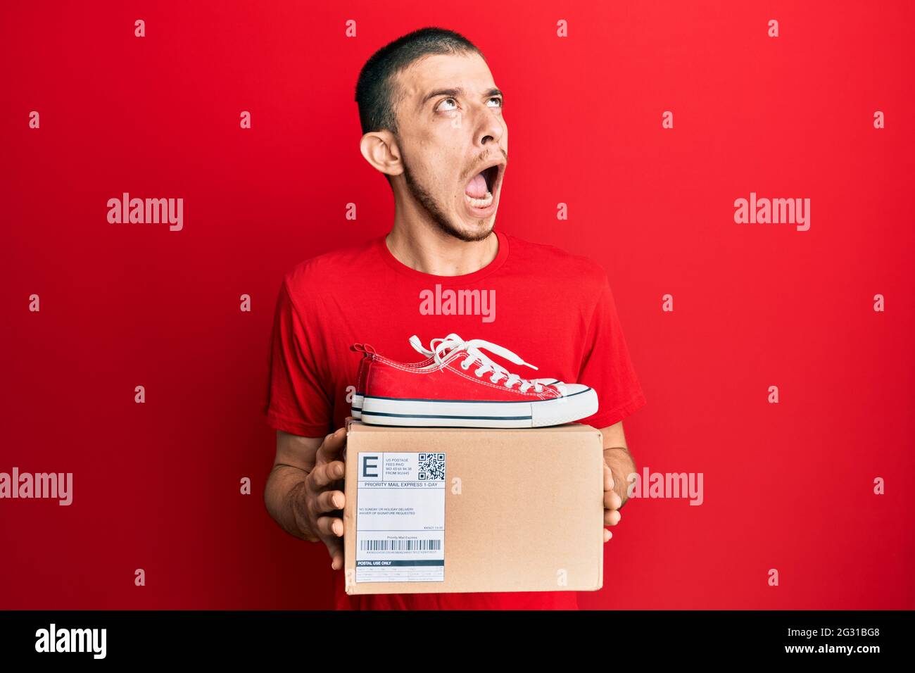 Hispanic young man holding casual red shoes and package angry and mad screaming frustrated and ...