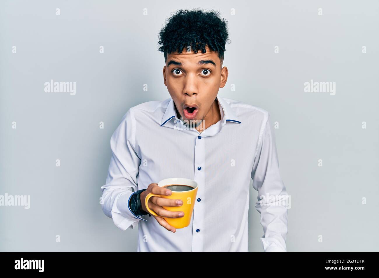 Young african american man drinking a cup of coffee scared and amazed with open mouth for ...