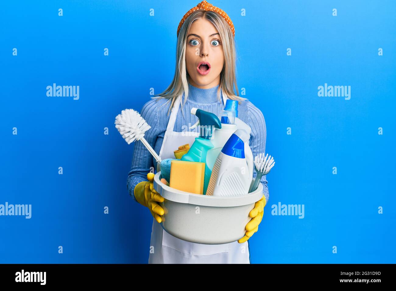 Beautiful blonde woman wearing cleaner apron holding cleaning products afraid and shocked with ...