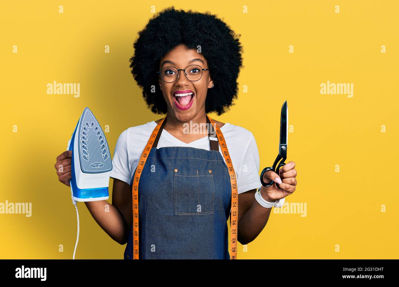 Young african american woman dressmaker designer wearing atelier apron holding iron and scissors ...