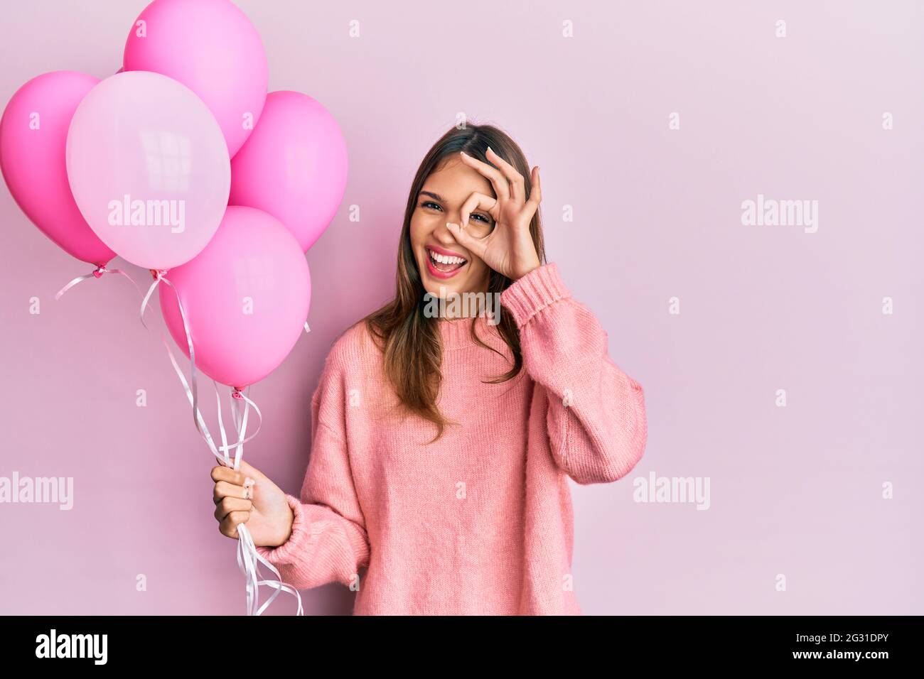 Young brunette woman holding pink balloons smiling happy doing ok sign ...