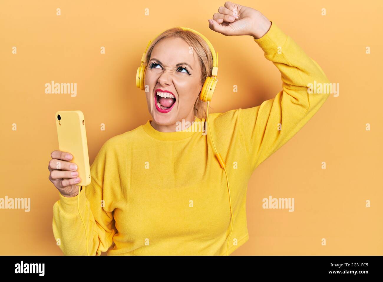 Young blonde woman dancing wearing headphones angry and mad screaming ...