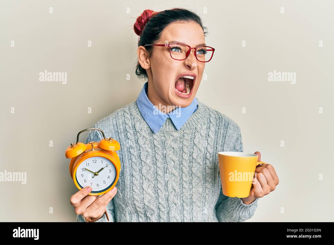 Young caucasian woman holding alarm clock drinking coffee angry and mad ...
