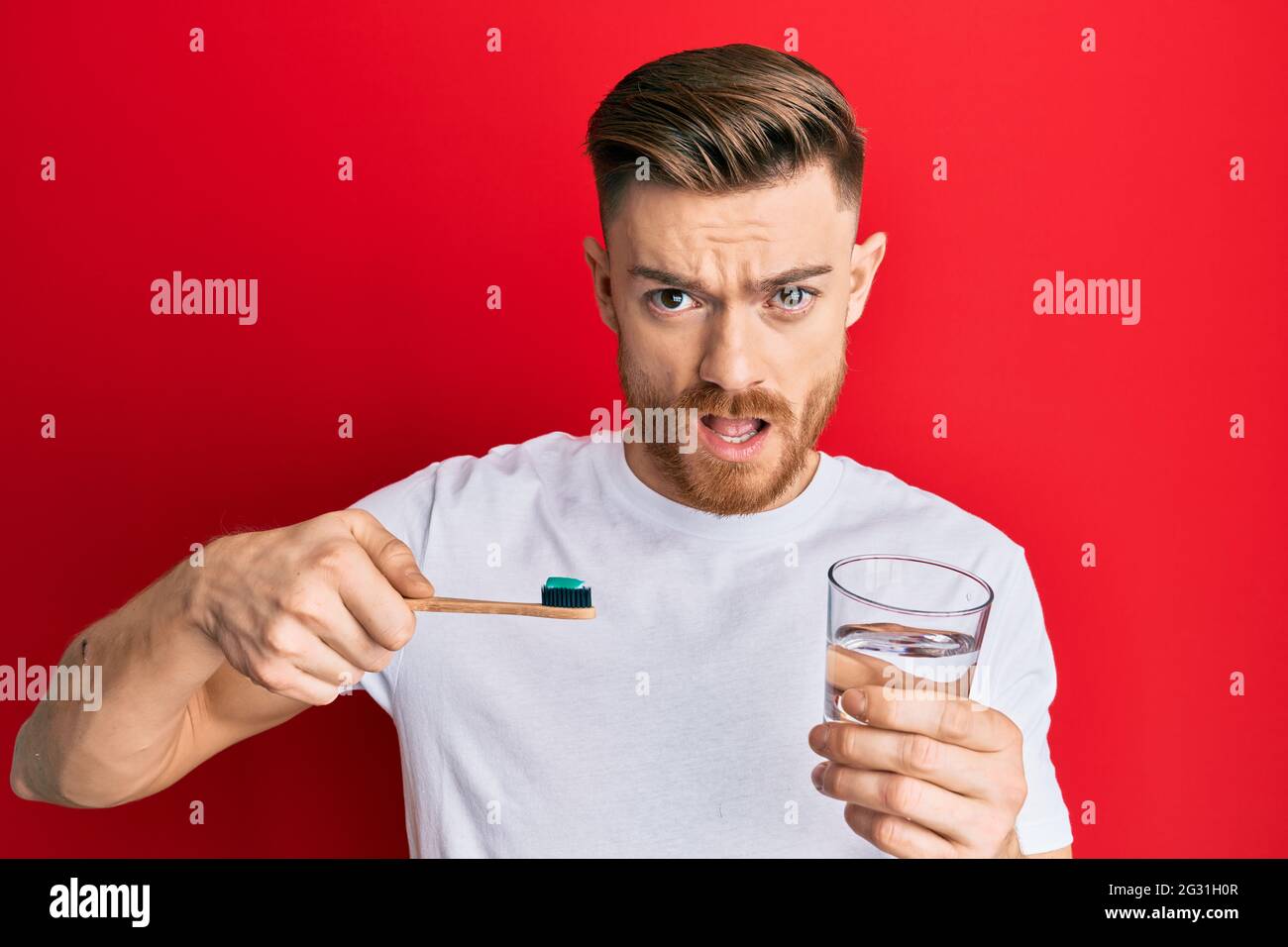 Young redhead man holding toothbrush with toothpaste and glass of water ...