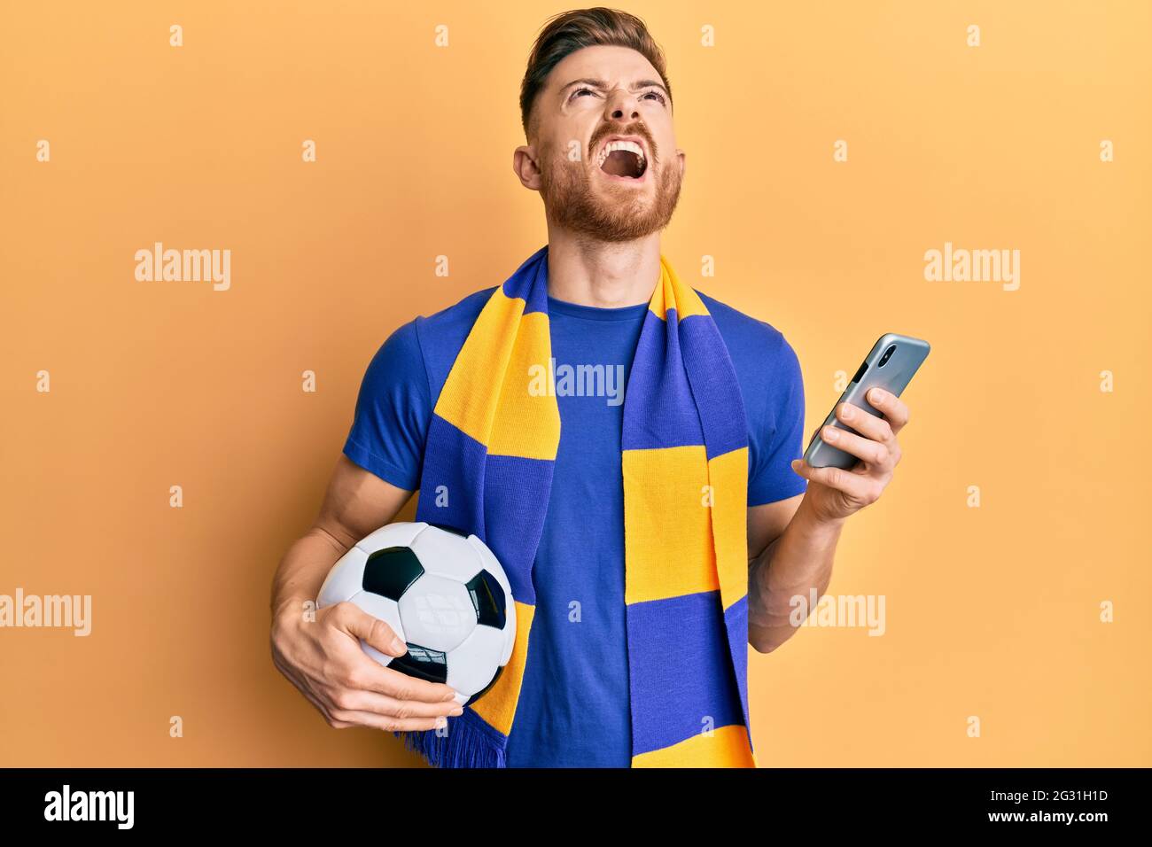 Young redhead man wearing hooligan scarf and using smartphone angry and ...