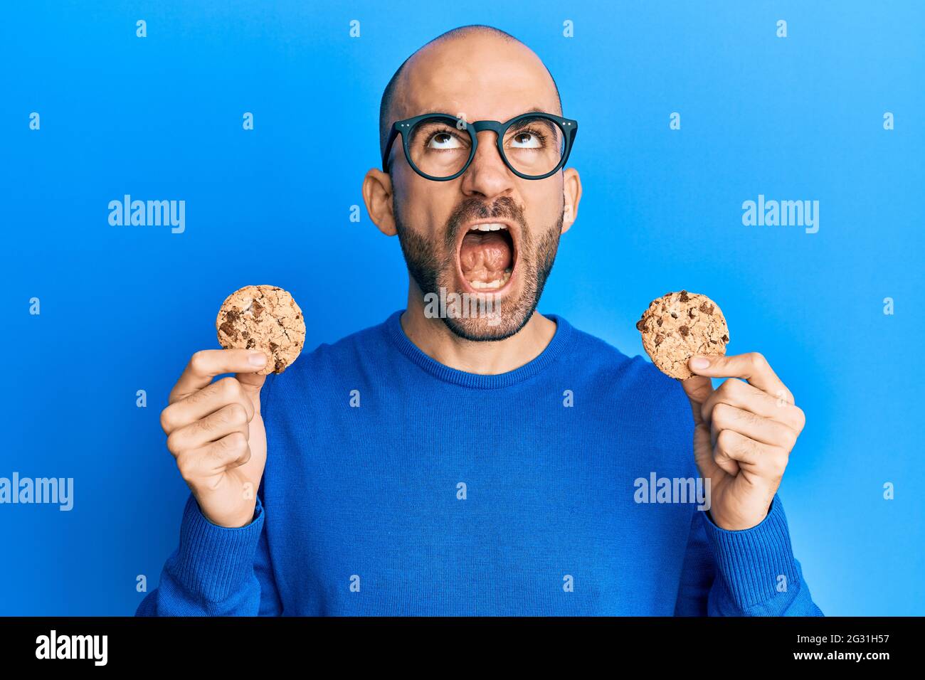 Young hispanic man holding chocolate chips cookies angry and mad ...