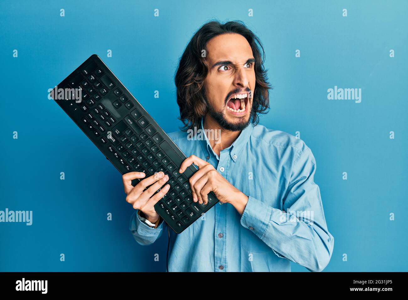 Young hispanic man holding keyboard angry and mad screaming frustrated ...