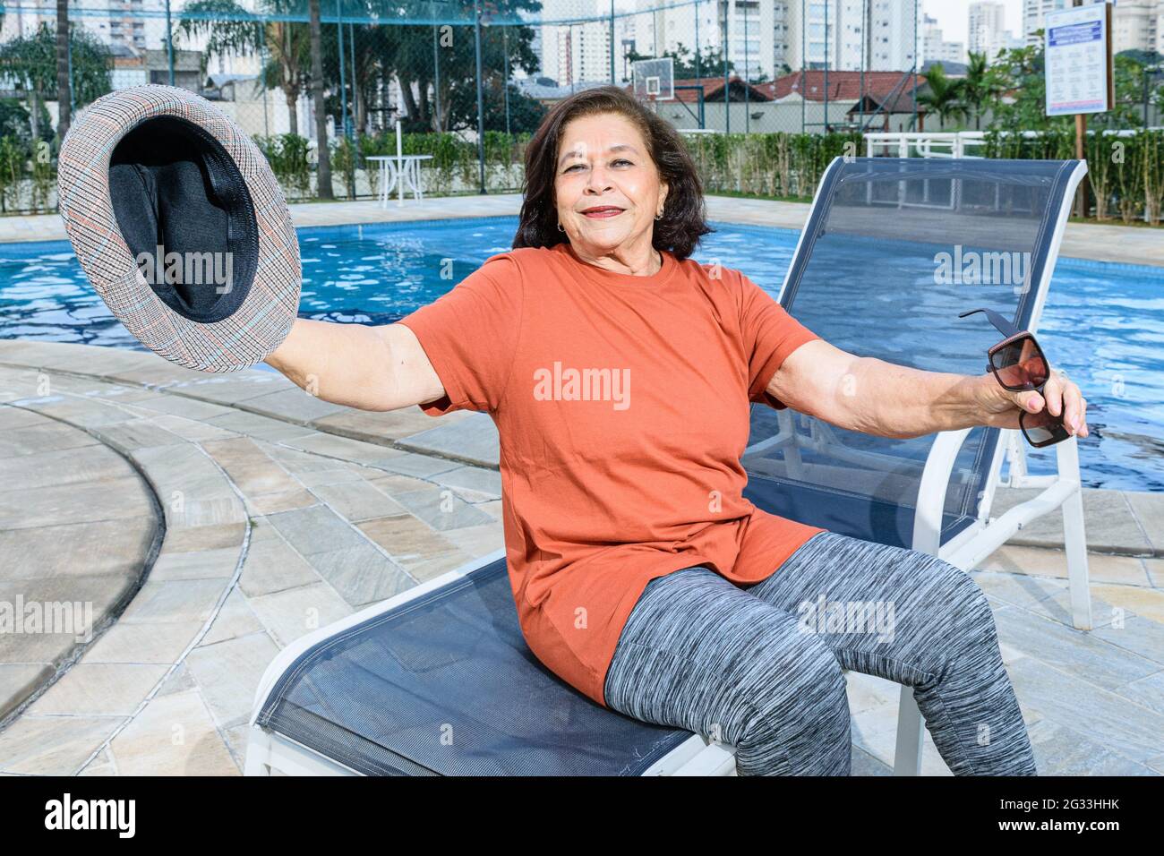 74-year-old elderly woman standing by the pool, hat and glasses in hand ...