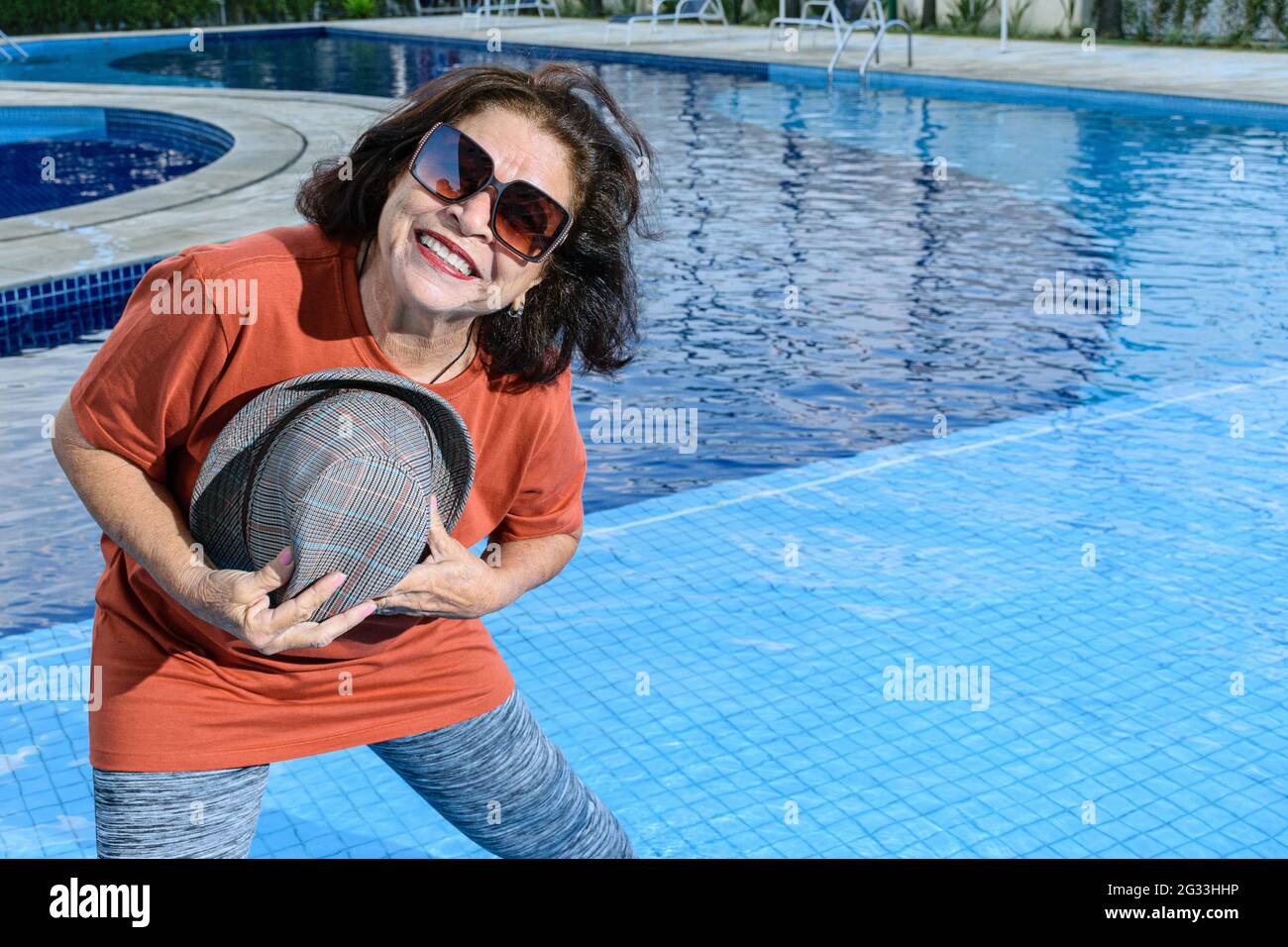 74 years old woman, with big smile, in swimming pool and holding hat ...