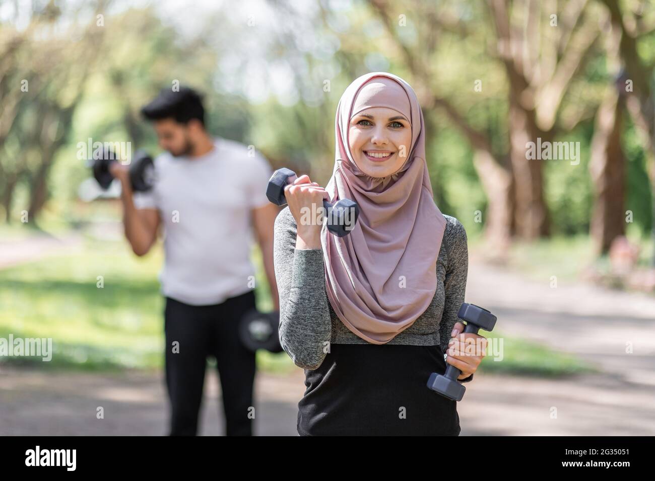 Portrait of smiling woman in hijab and sport clothes using dumbbells ...