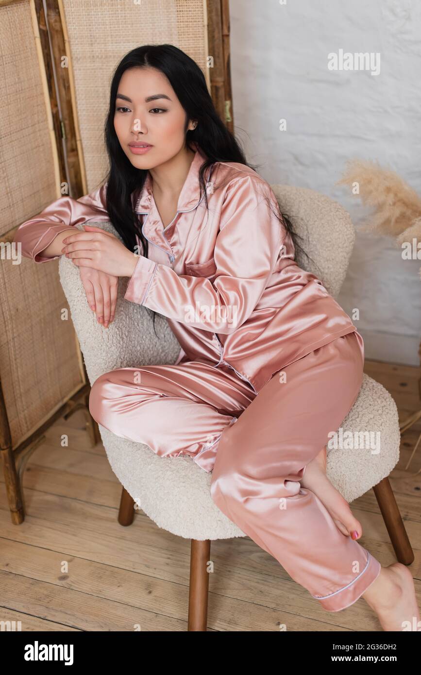 Dreamy Young Asian Woman In Pink Silk Pajamas Sitting On Chair In