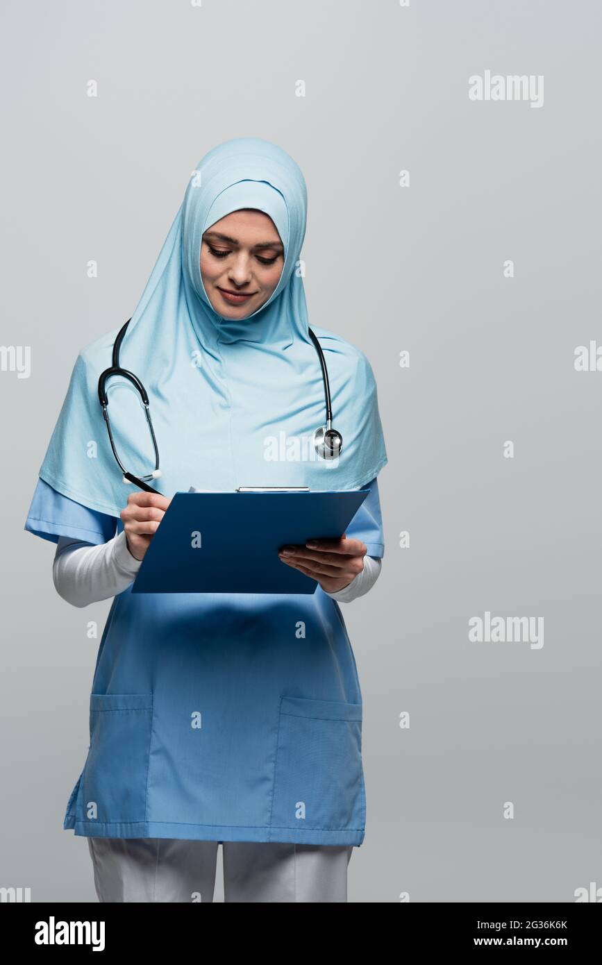 pretty muslim doctor in hijab and uniform writing on clipboard isolated ...
