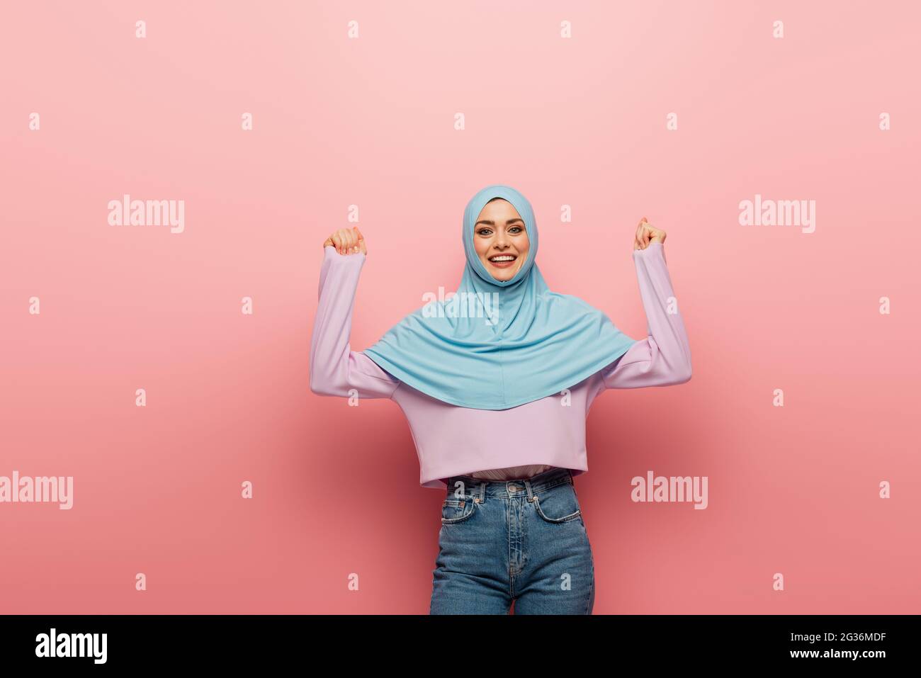 excited muslim woman in hijab and jeans showing rejoice gesture on pink ...