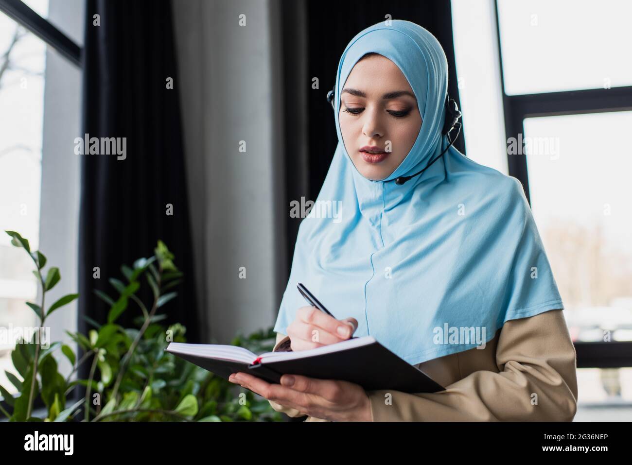 young muslim woman writing in notebook while working in call center ...