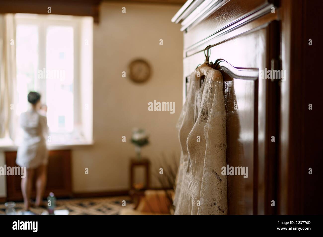 A bride in a robe stands at the window in a hotel room, a wedding dress ...