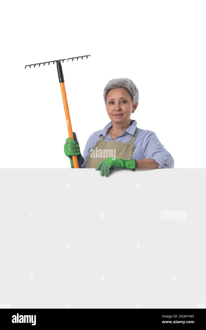 Mature asian woman gardener with rake standing behind blank banner with ...