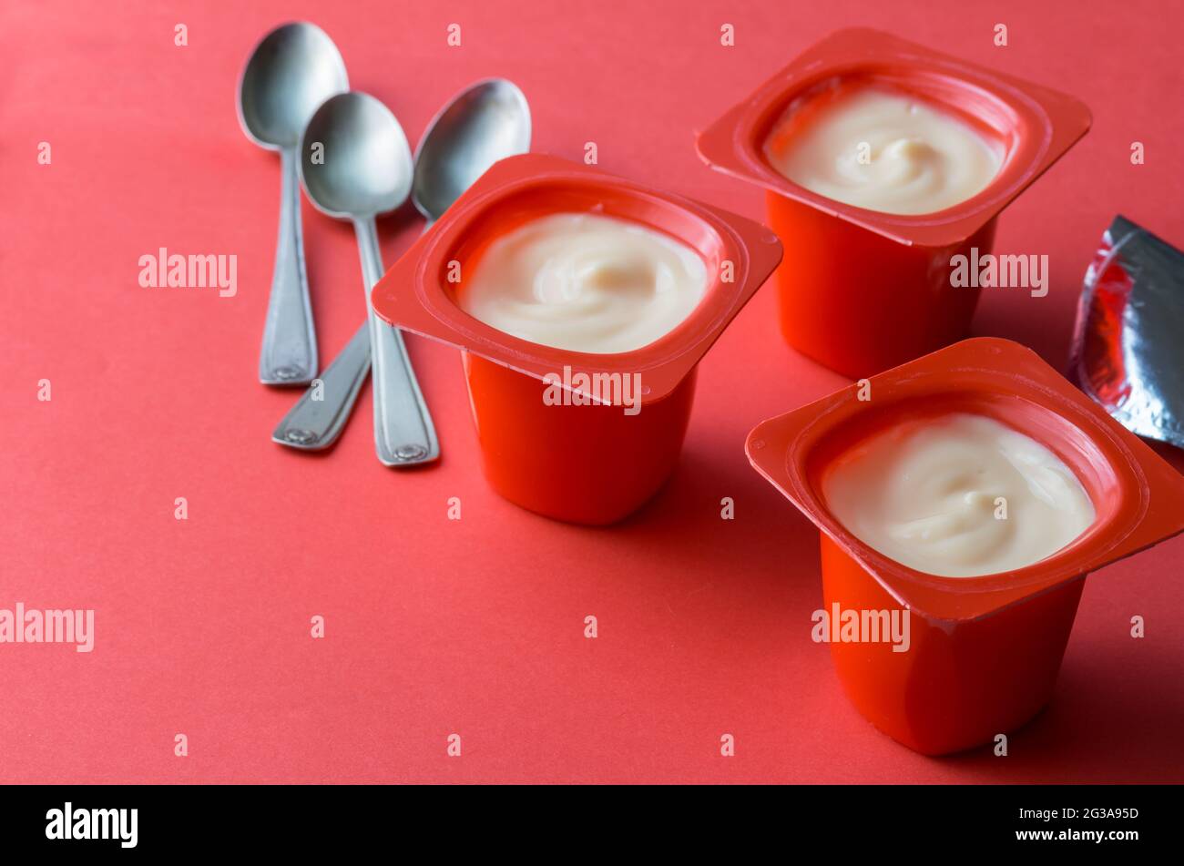 Three red yogurt plastic pots with spoons and foil on red background ...
