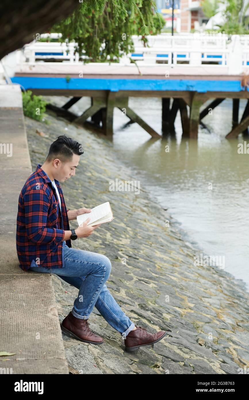 Serious young Vietnamese student in casual outfit sitting at riverside ...