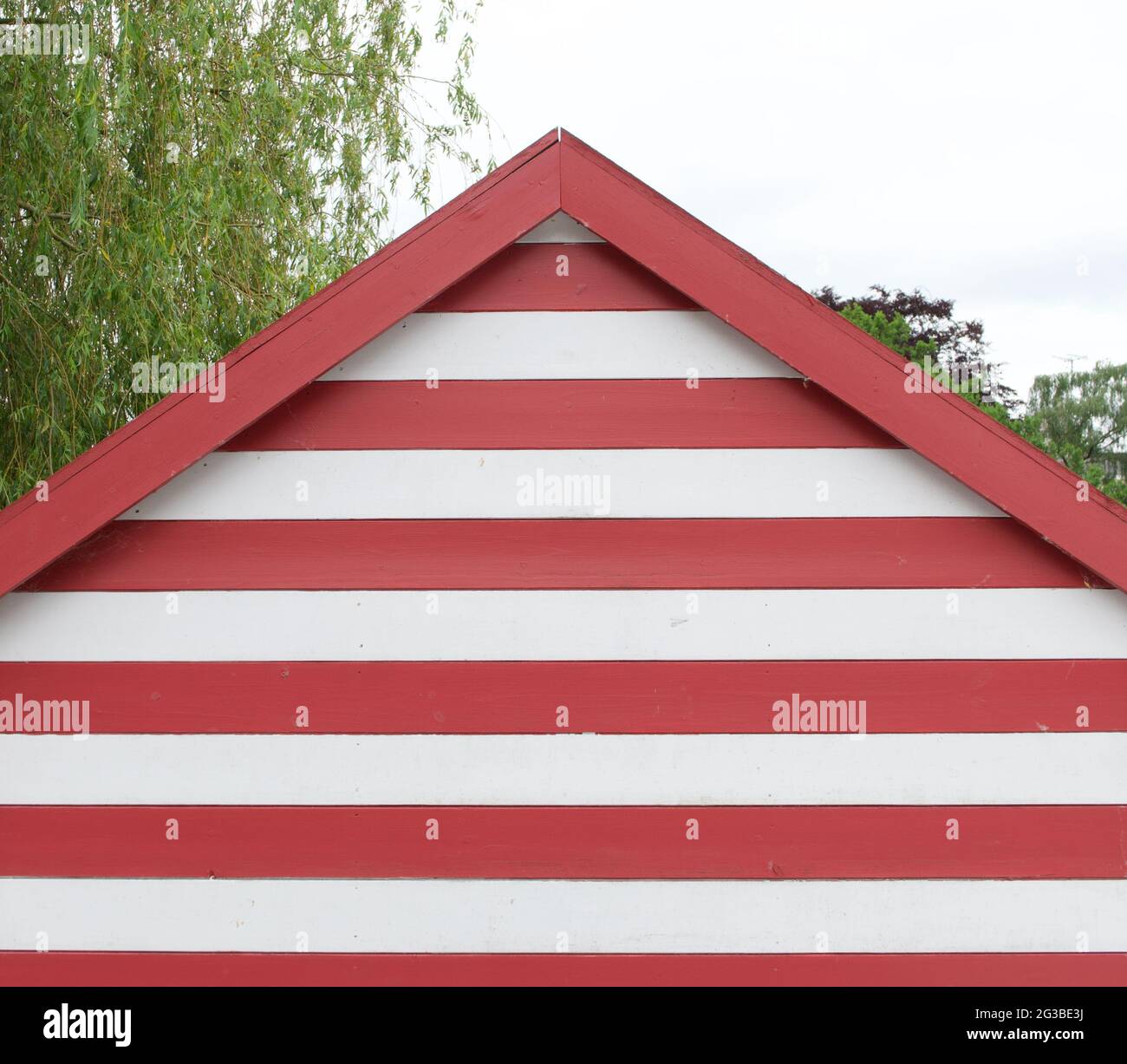 Old English beach hut painted in red and white stripes beside trees ...