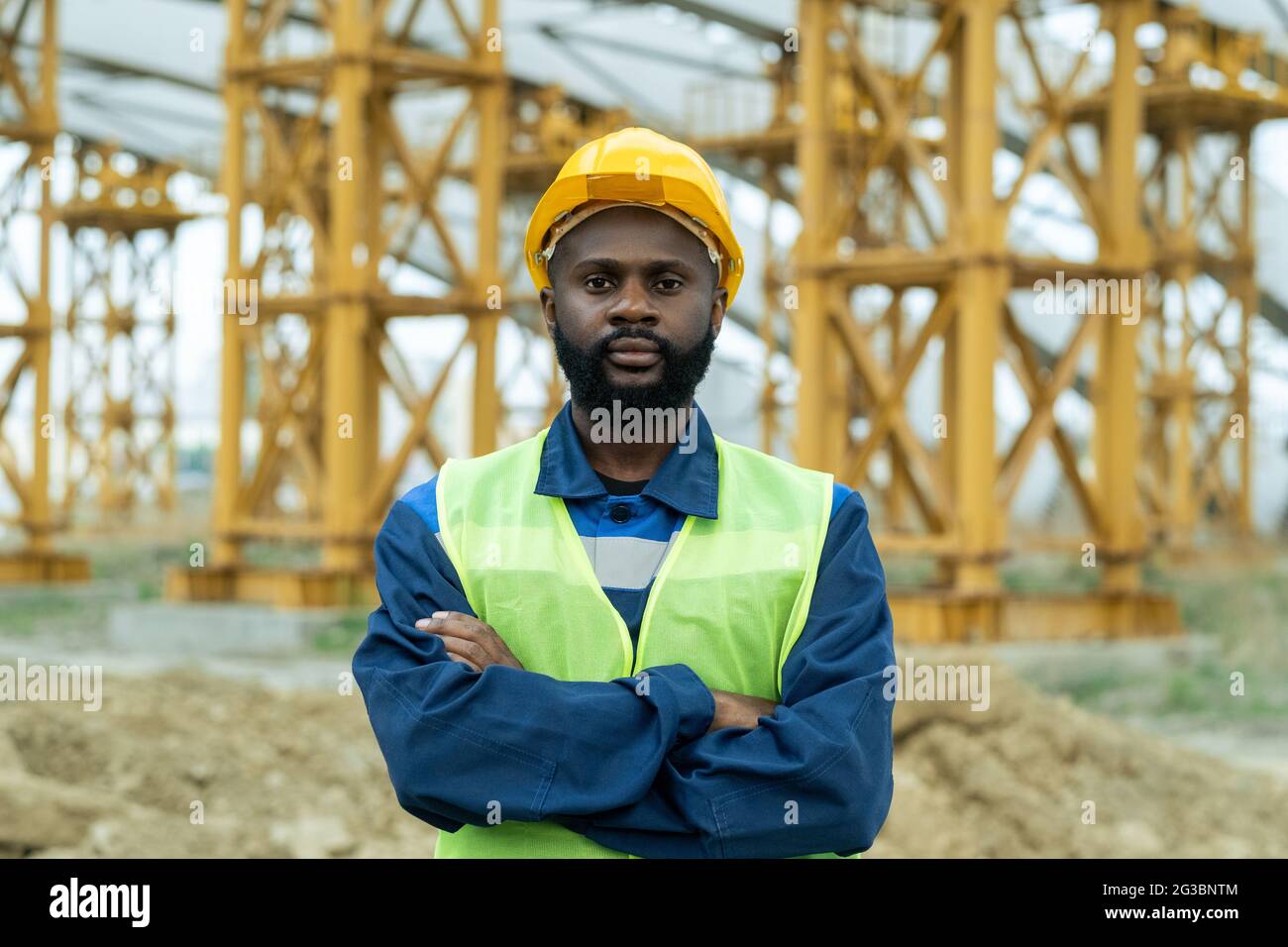 Young African construction worker in uniform and hardhat crossing arms ...