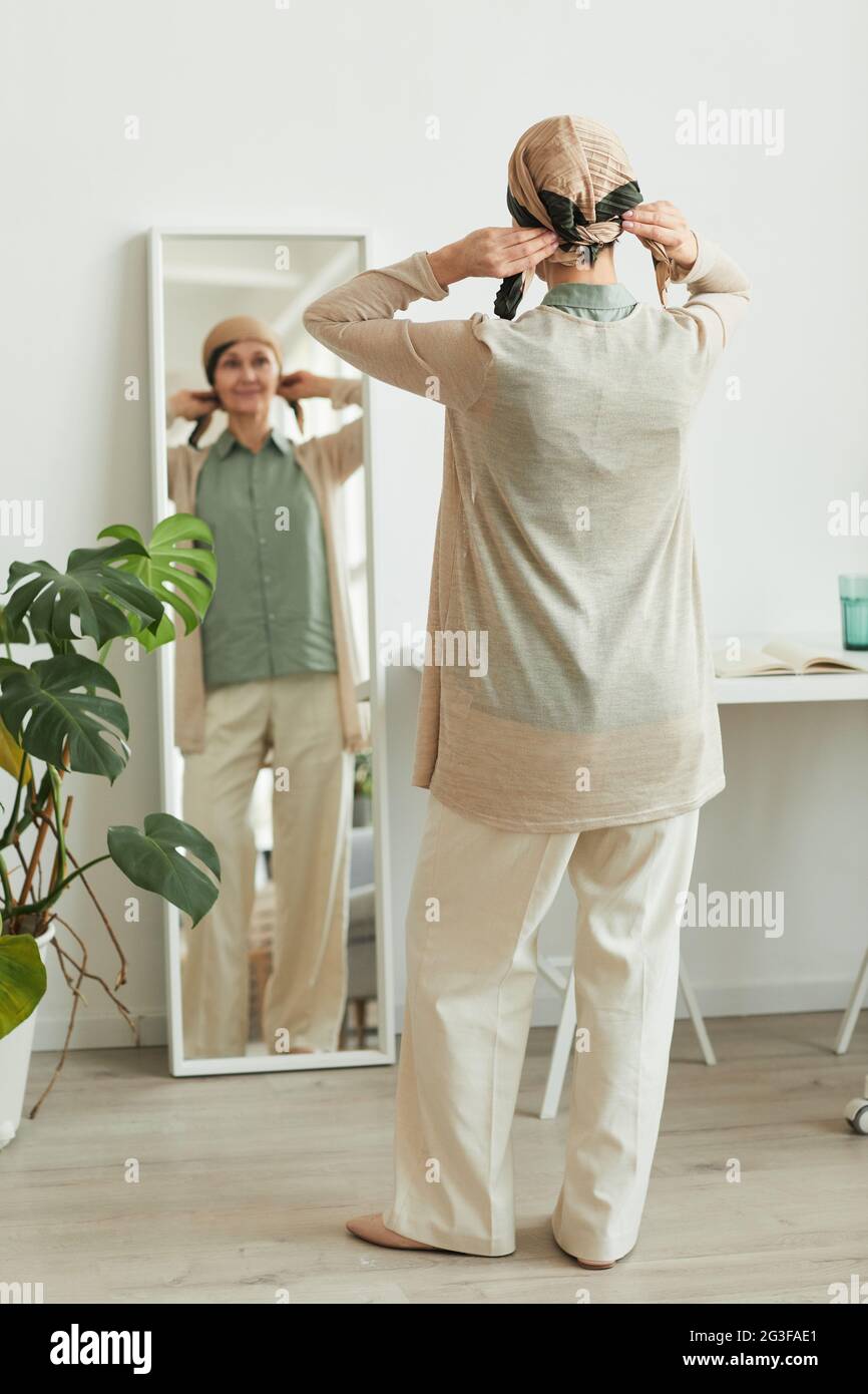 Full length back view at mature woman putting on headscarf by mirror ...