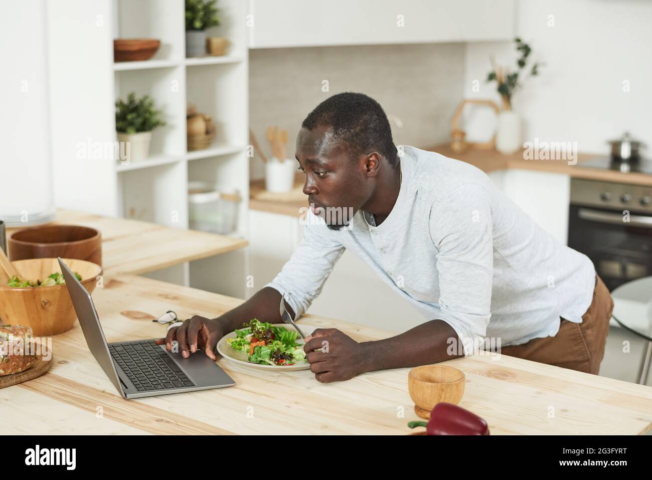 Young man eating vegetable salad and using laptop in the kitchen Stock ...