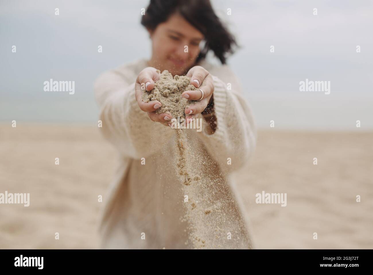 Stylish young woman in sweater releasing sand on beach, hands close up ...