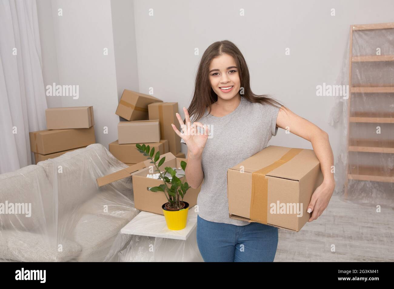 Pretty young woman standing with cardboard box in her hand showing ok ...