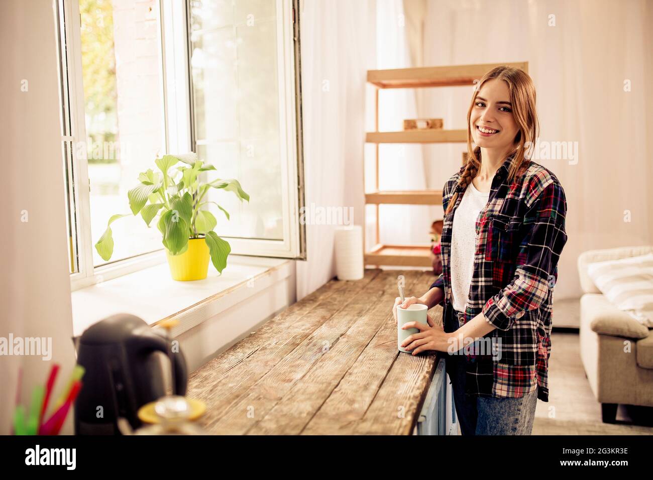 Attractive young girl standing close to window with cup of coffee or ...