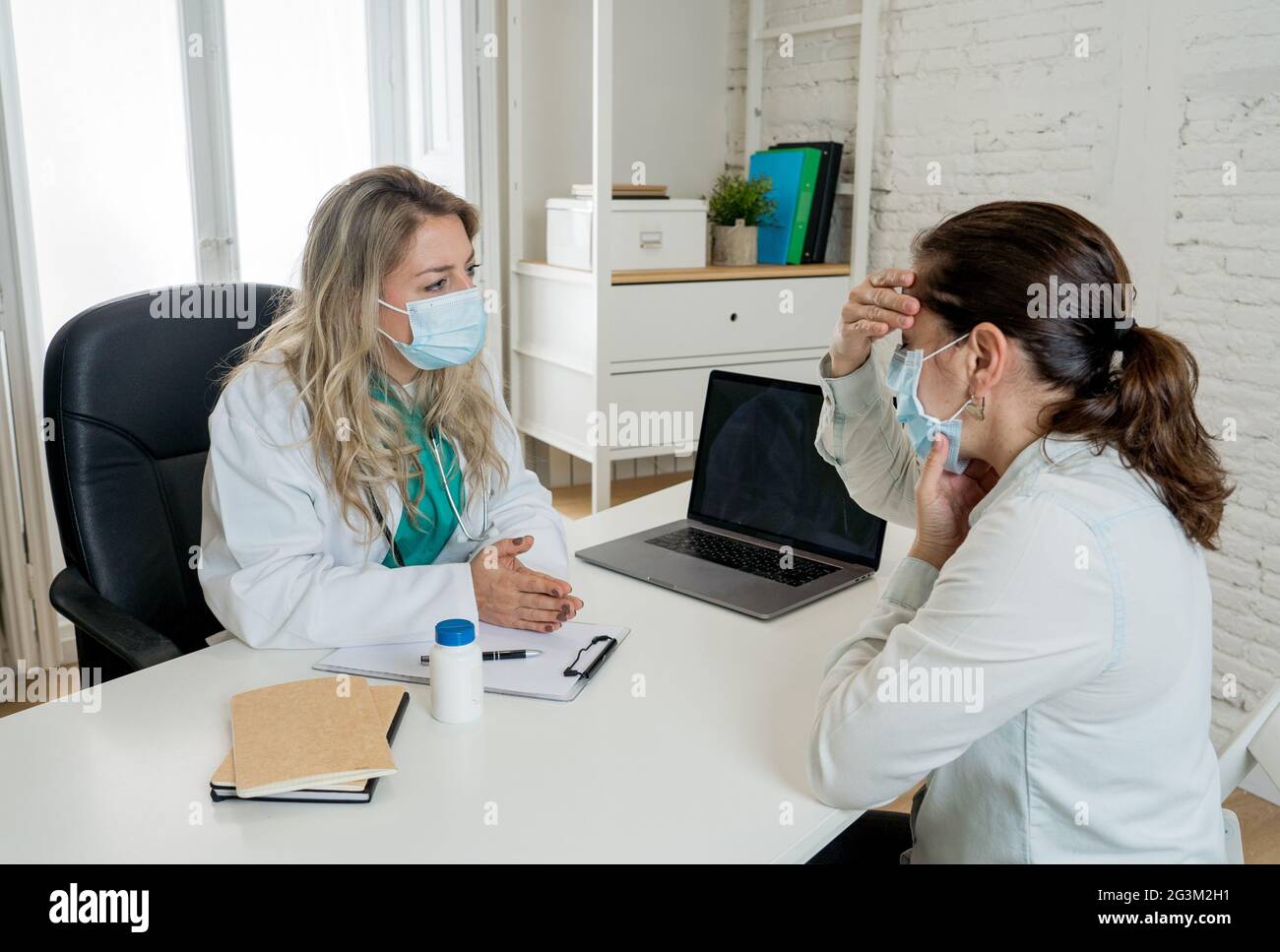Female doctor and woman patient with face masks talking in hospital o ...