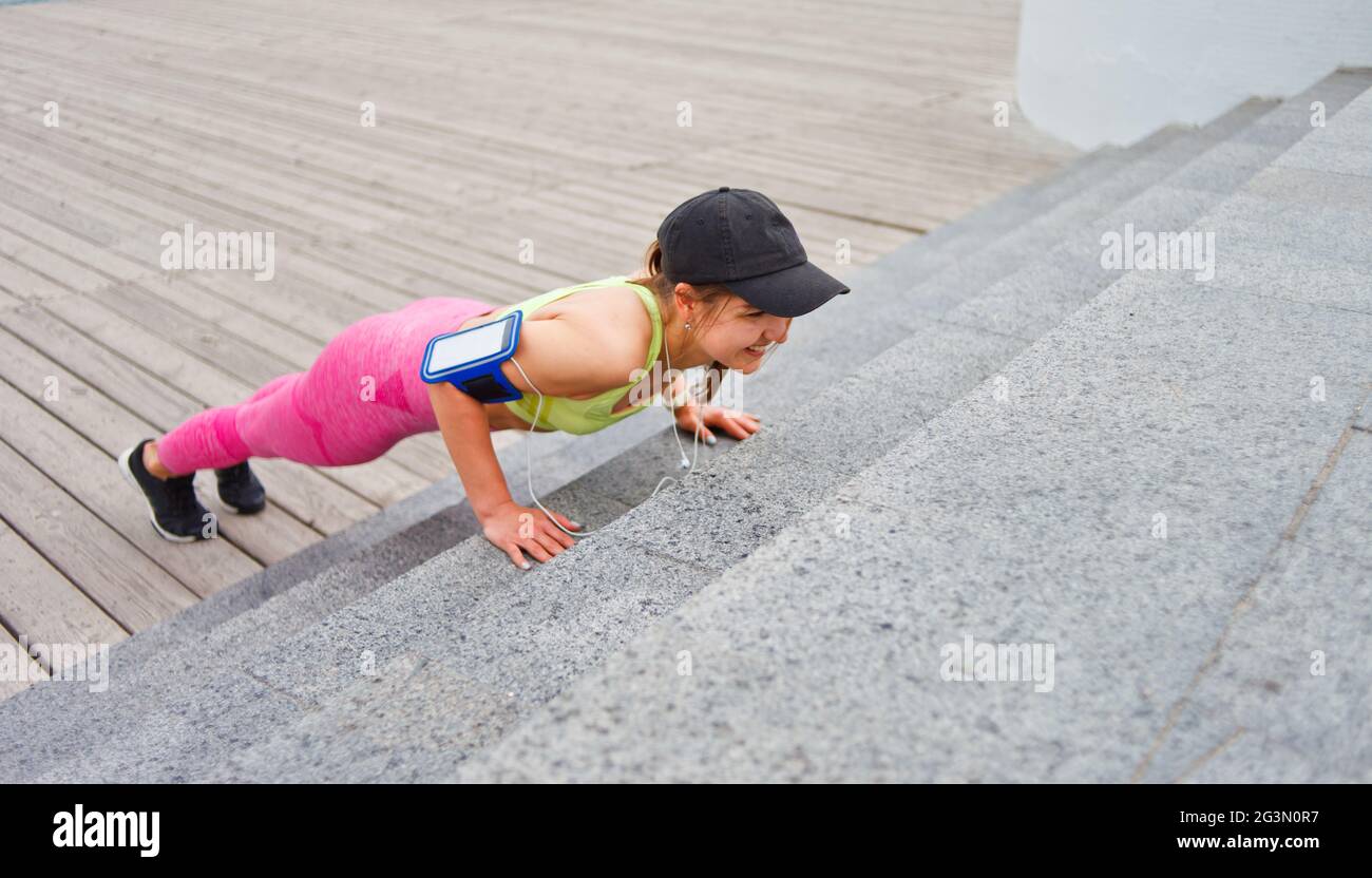 Cheerful fit woman in sportswear and cap doing push-ups from the stairs ...