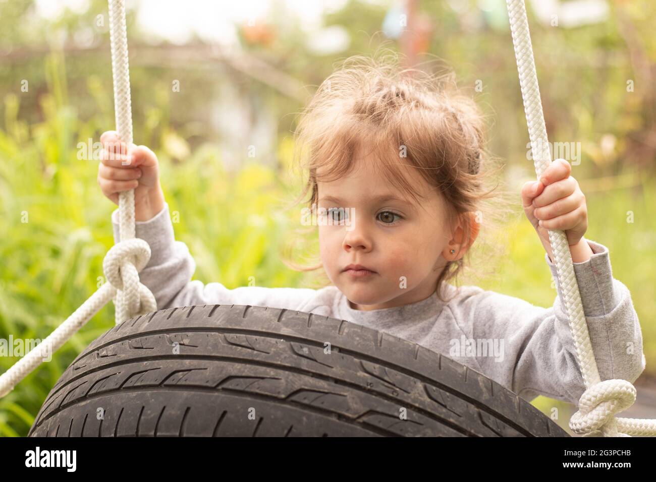 little cute red-haired girl riding a black wheel swing tied with ropes ...