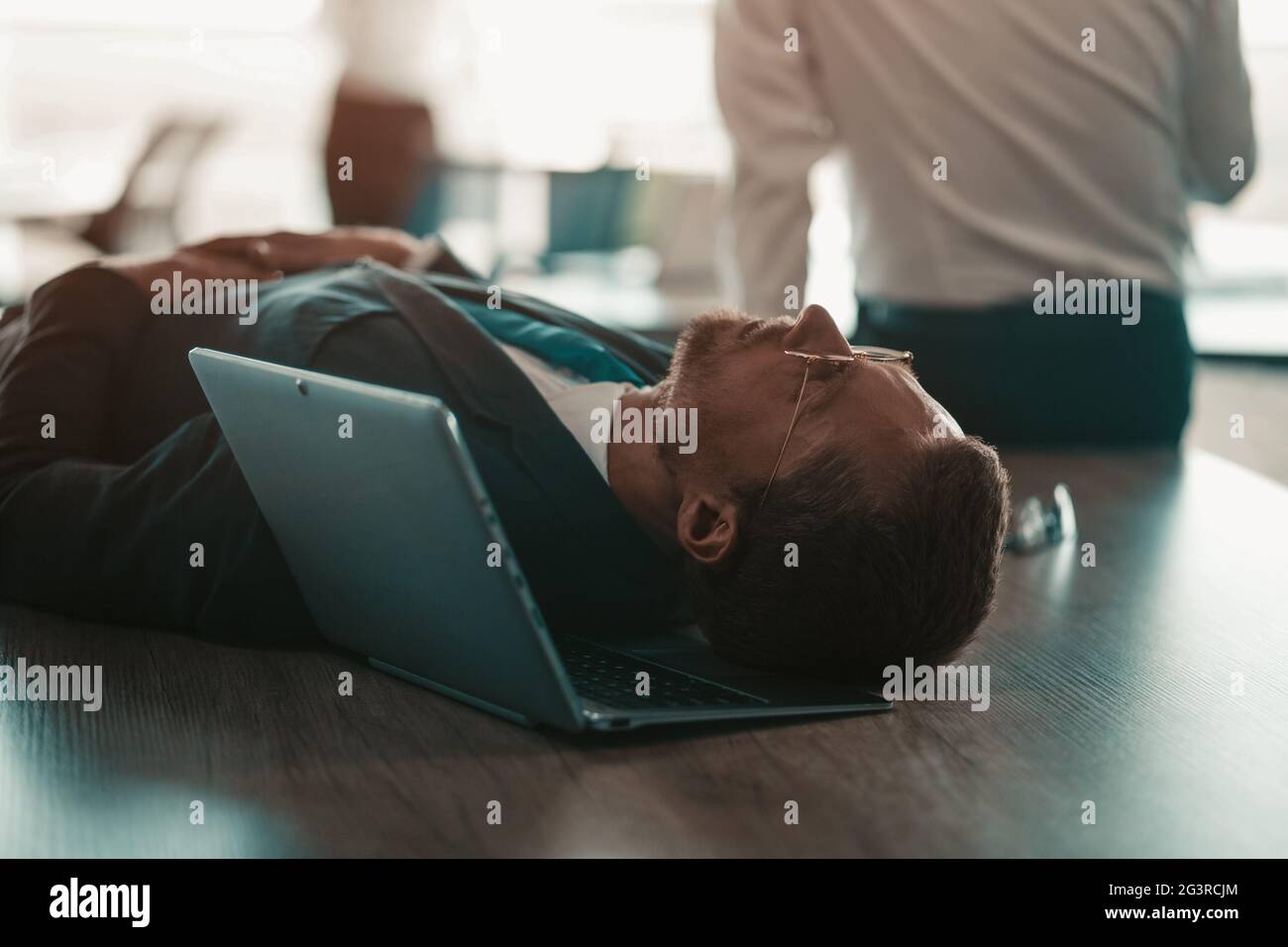Tired office worker is lying on his desktop on his laptop. He's wearing ...