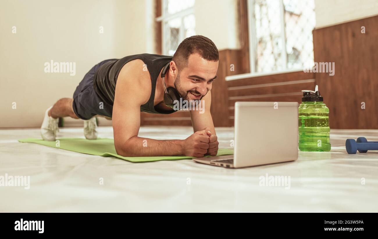 Fitness trainer doing plank on the mat pad with laptop doing online ...