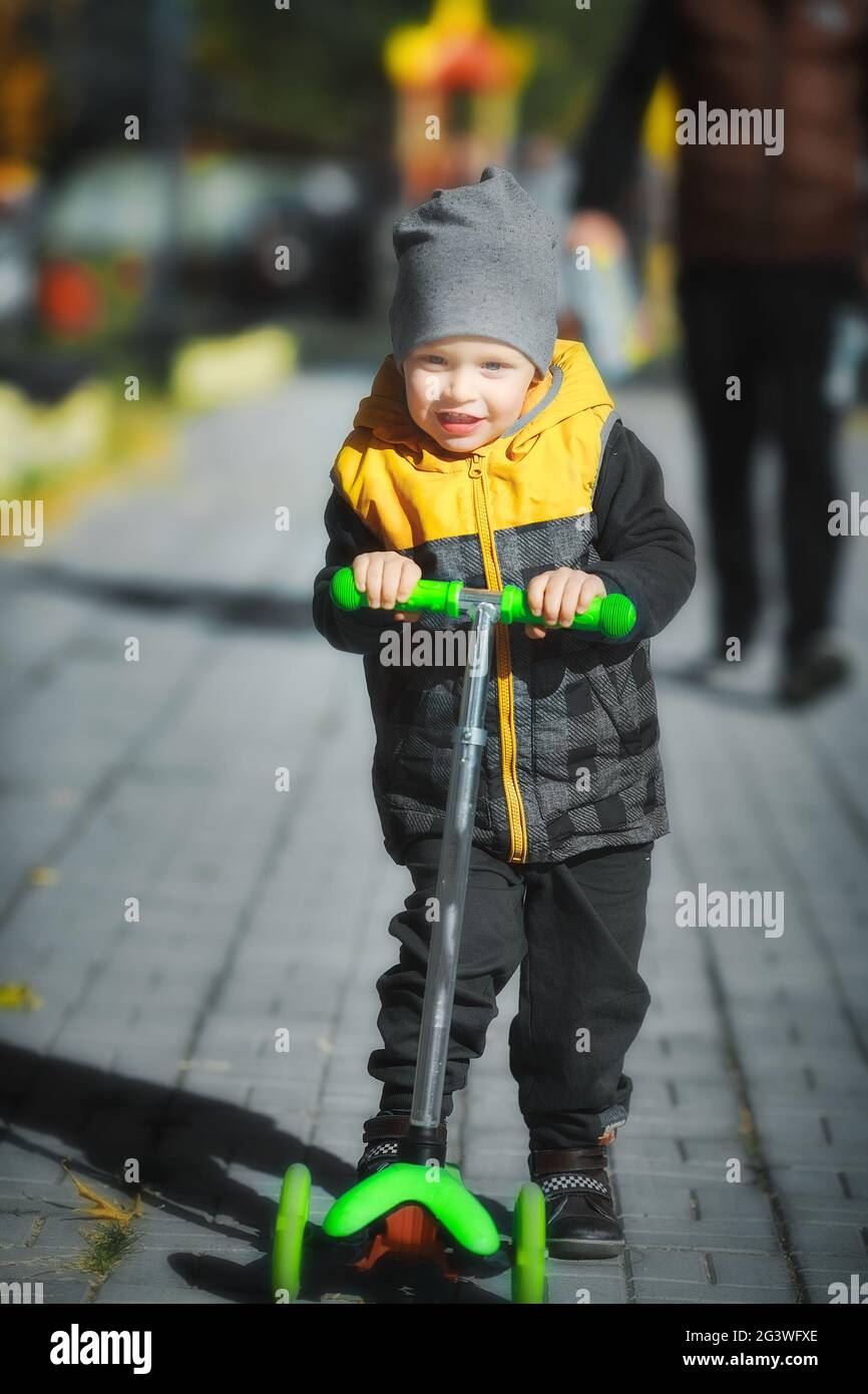 Happy and emotional toddler rides a three-wheeled scooter in the park ...