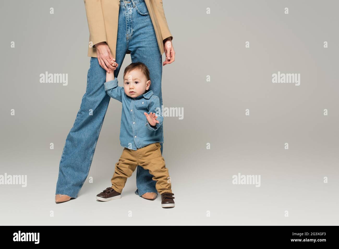 cropped view of mother supporting and holding hand of baby boy on grey ...