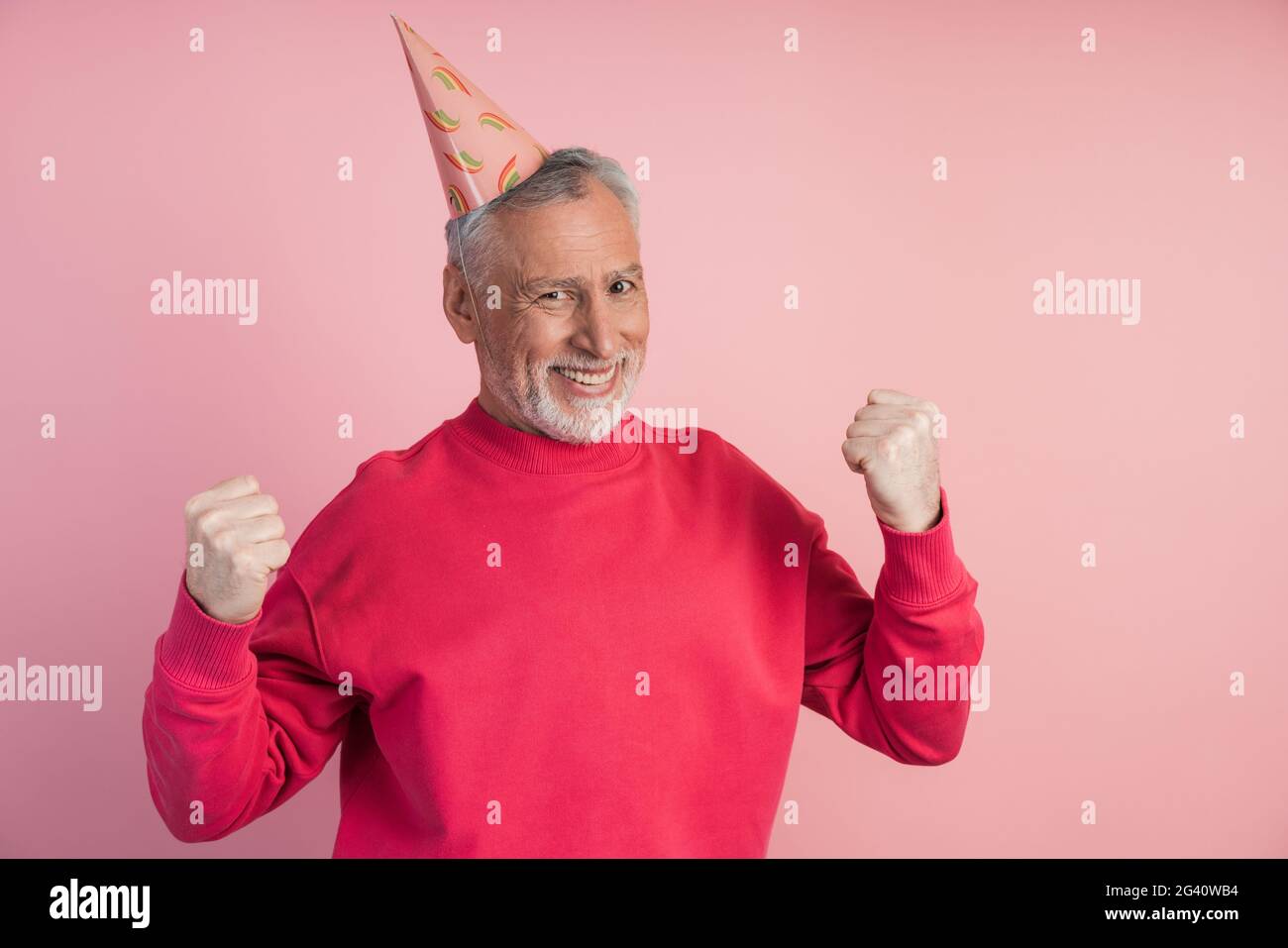 Confident senior man in a holiday hat rejoices in victory. Man isolated on pink background, copy ...