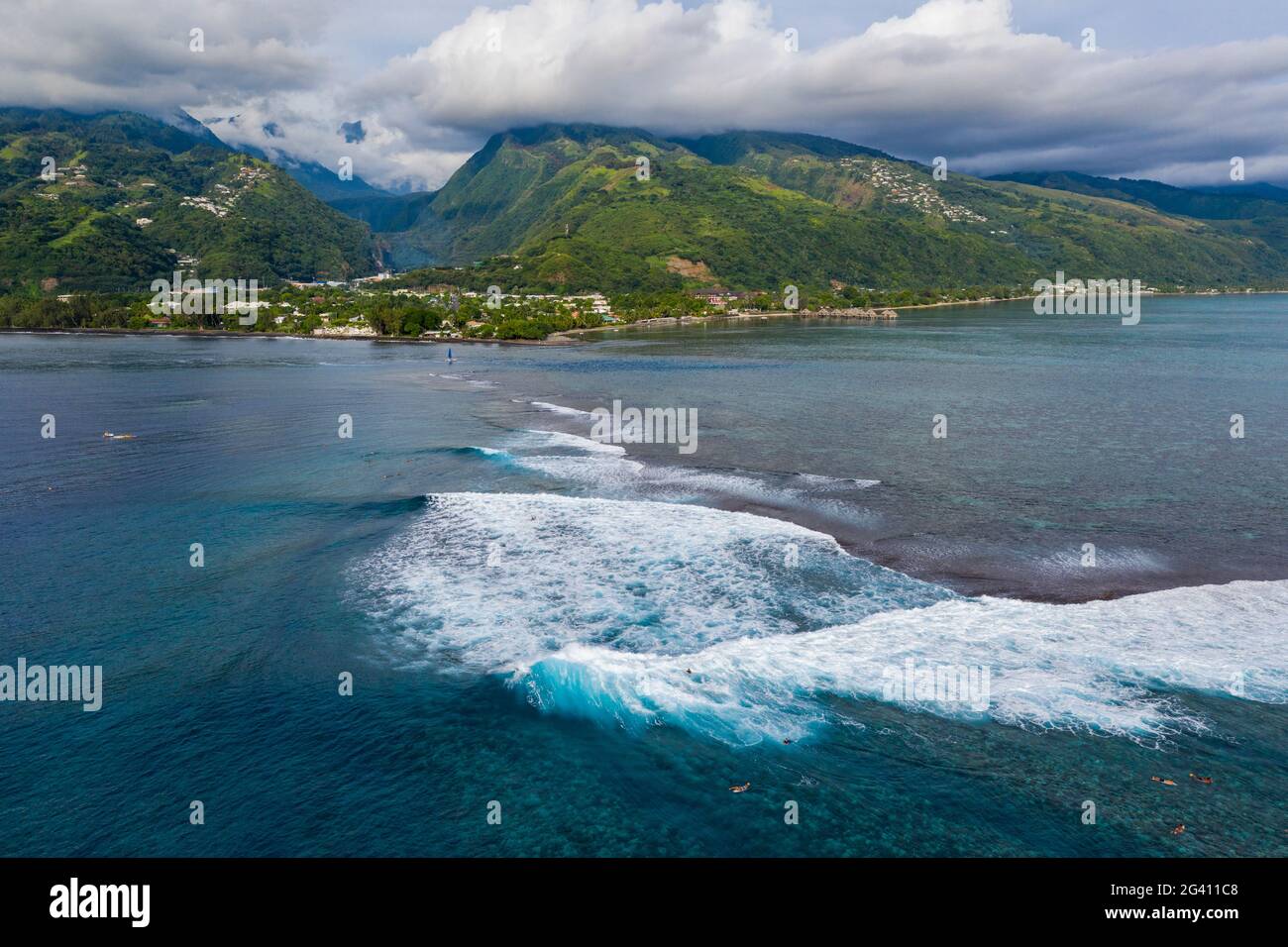 Aerial view of surfers on wave on reef with coast and mountains behind ...