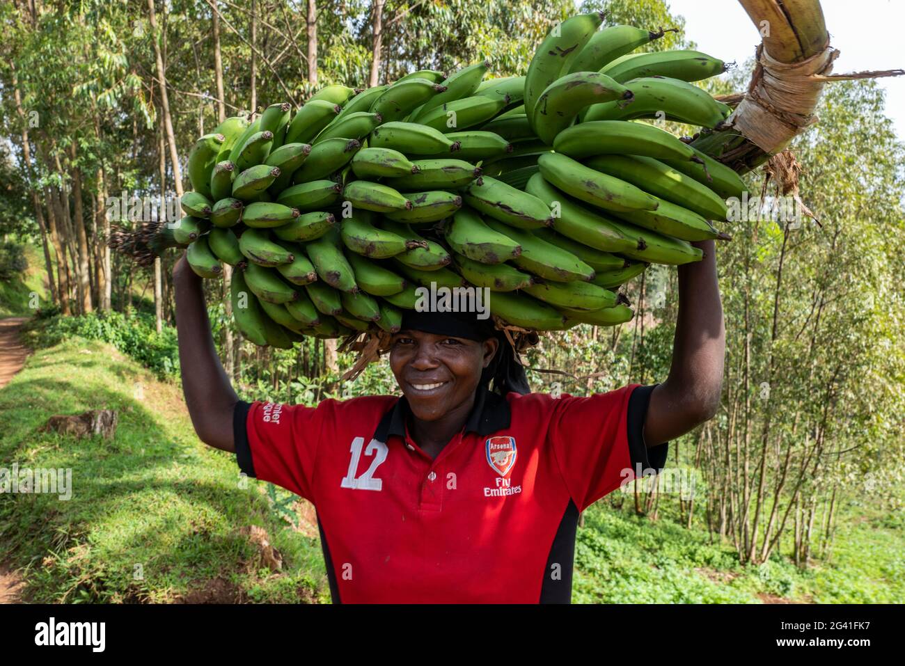 Smiling woman in Arsenal soccer jersey carries heavy banana tree on ...