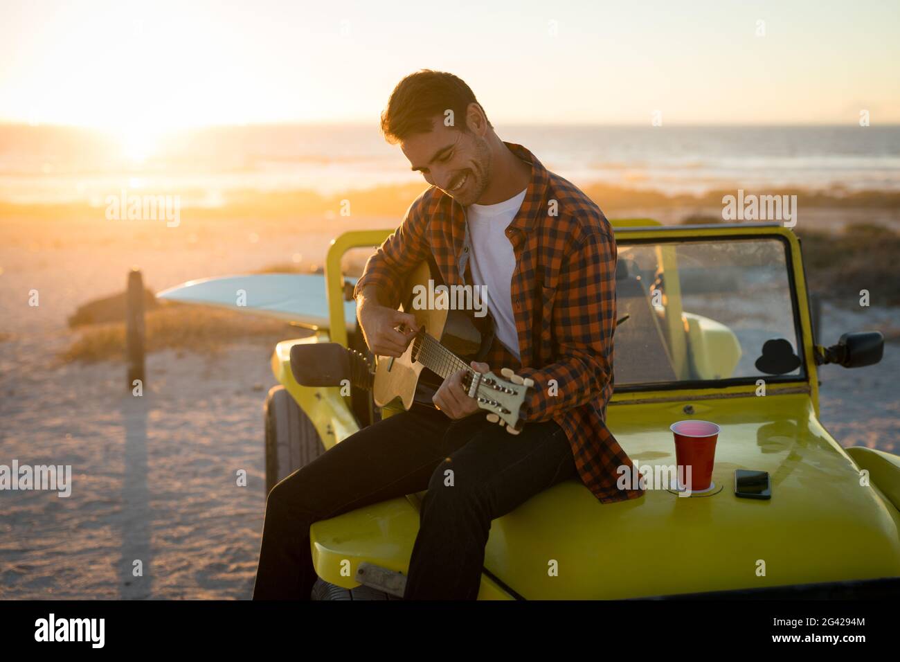 Happy caucasian man sitting on beach buggy by the sea playing guitar ...