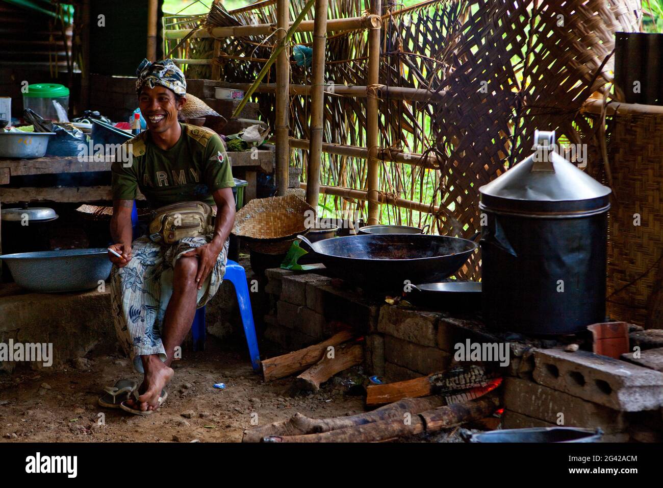 A Balinese cook, sitting in a ramshackle kitchen in a hut, smoking and ...