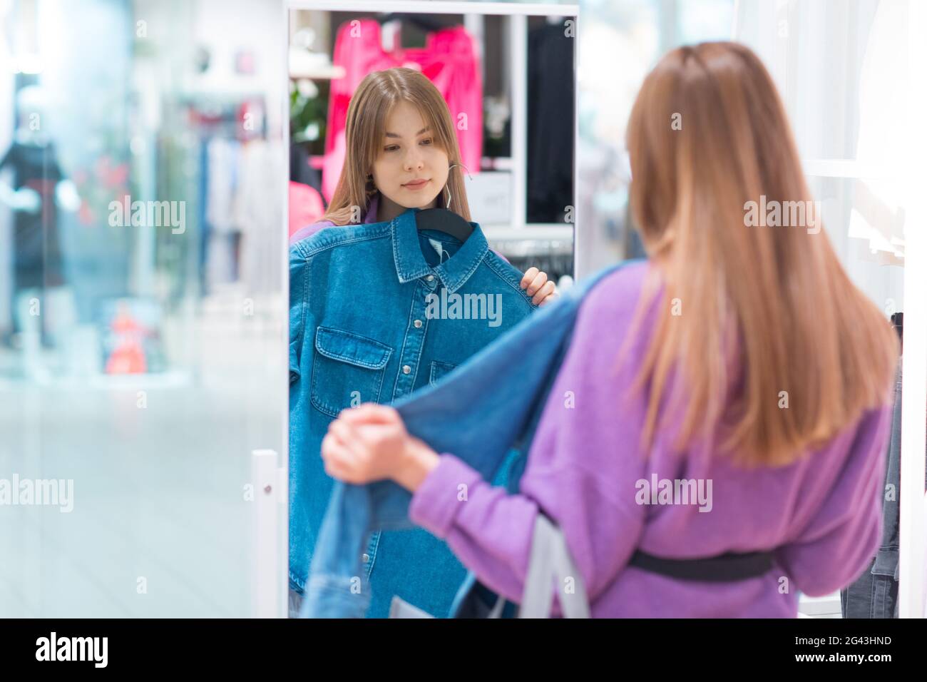 Young woman chooses and trying on and looking mirror clothes in shop ...