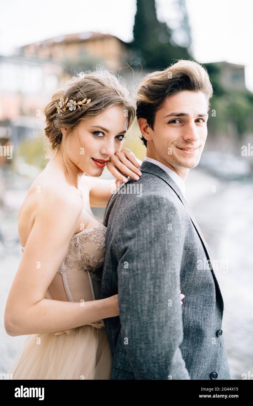 Smiling bride hugs groom from behind while standing on the promenade of ...