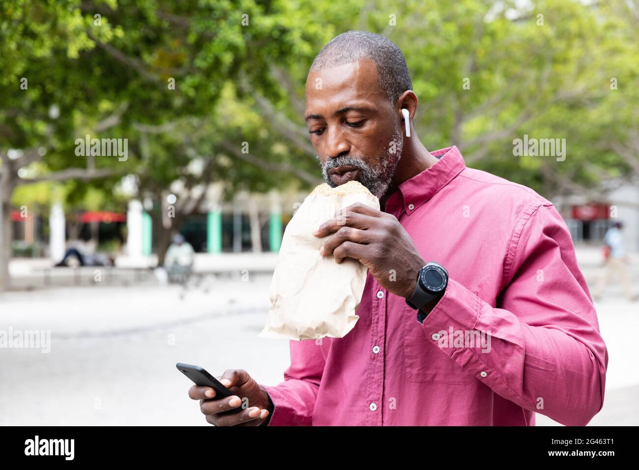 African American man using his phone and eating a takeaway sandwich ...