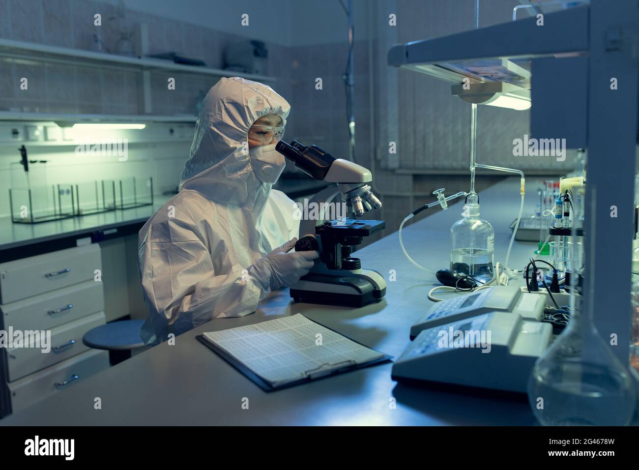 Young female scientist in coveralls looking in microscope by workplace ...