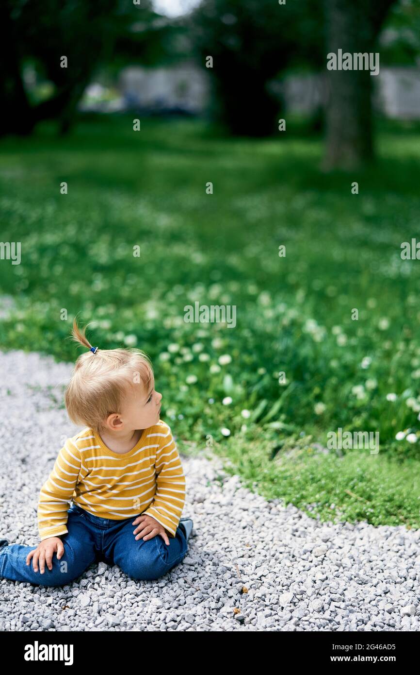 Little girl sits on a gravel path in a green park, turning her head to ...
