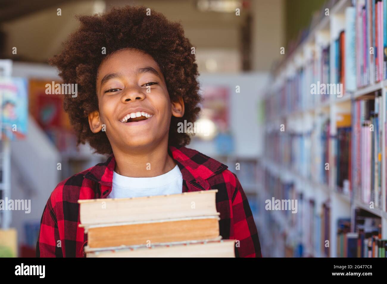 Laughing african american schoolboy carrying stack of books in school ...