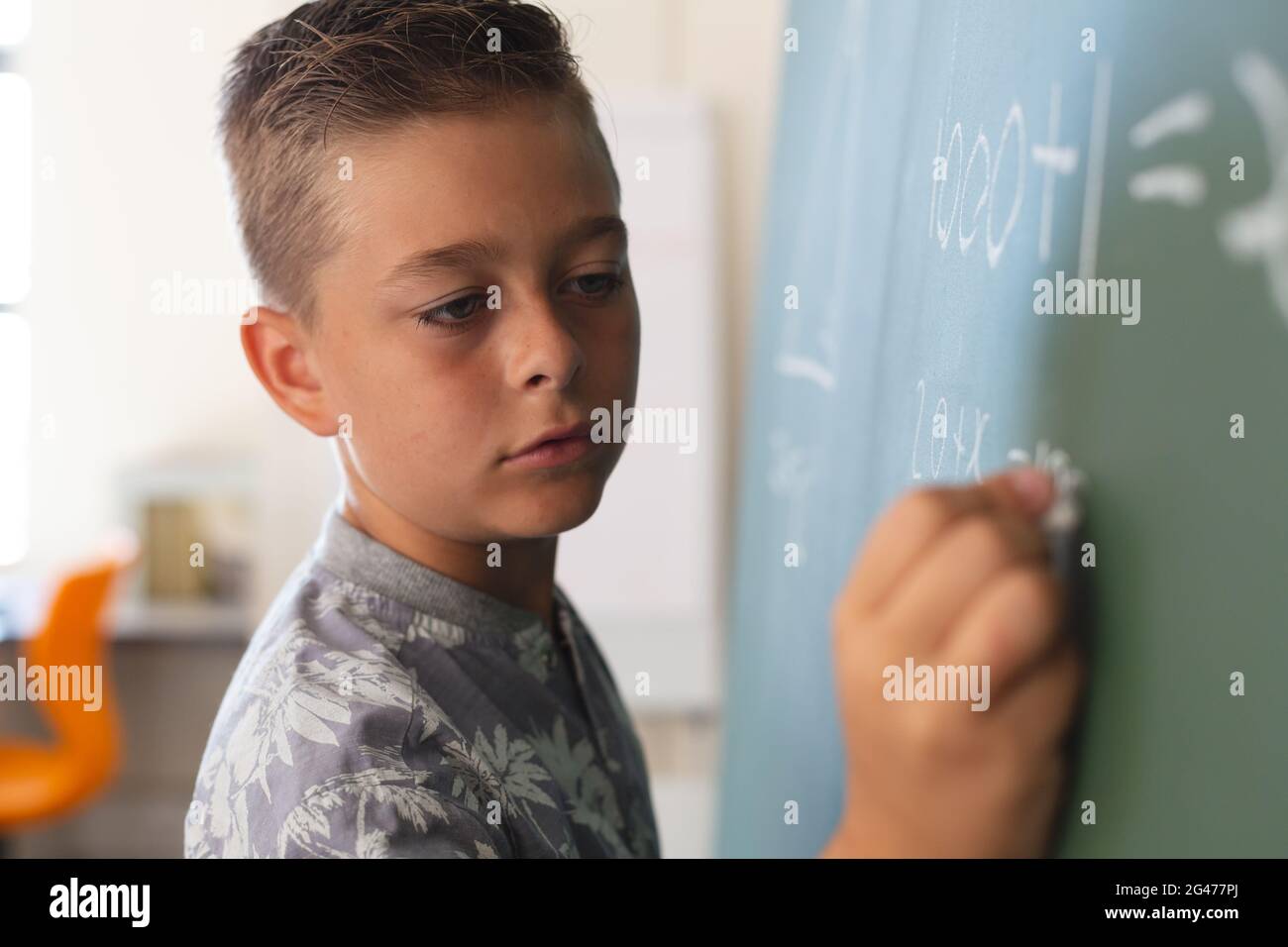 Caucasian boy standing at chalkboard writing in classroom during maths ...