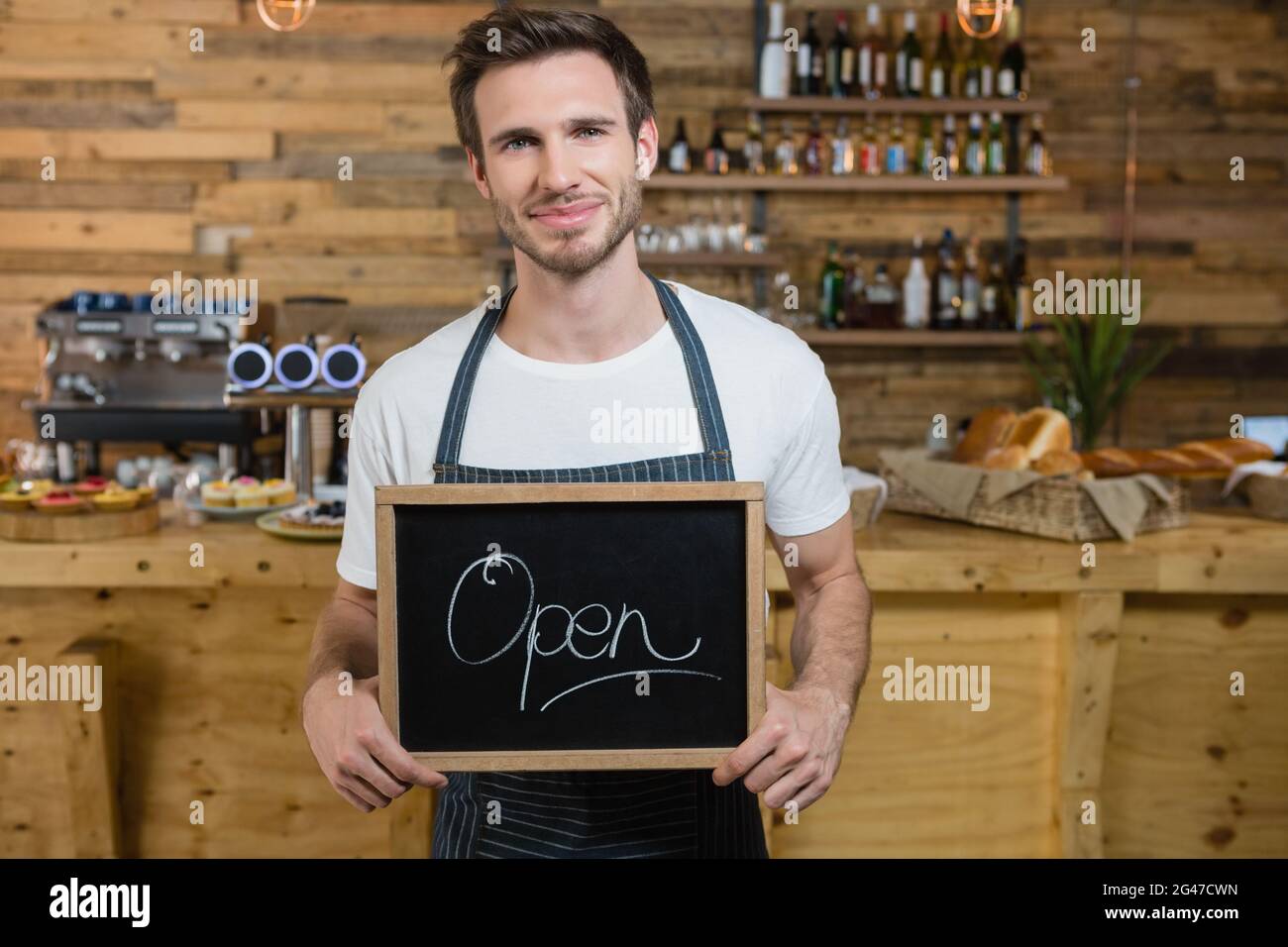 Portrait of smiling waiter standing with open signboard at counter ...